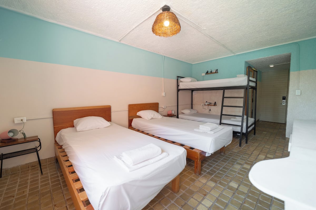 Quarto com duas camas de solteiro, uma cama beliche, mesa de cabeceira com luminária e banheiro com pia e espelho.