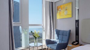 Quarto de hotel com uma janela grande, uma cadeira azul, uma mesinha de cabeceira com telefone e um quadro na parede.