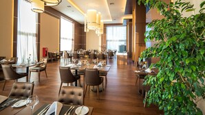 Um restaurante espaçoso com pé-direito alto, iluminação pendente moderna e uma grande planta de interior. As mesas estão dispostas com louça fina e copos de vinho.