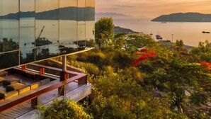 Um edifício moderno com fachada de vidro refletindo um pôr do sol sobre a água, rodeado por vegetação exuberante e barcos.