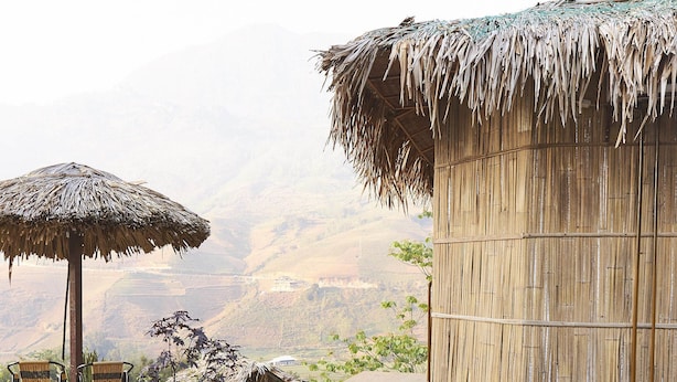 Uma área de estar rústica ao ar livre, com um toldo de palha, bancos de madeira e um caminho de pedra que leva a um bangalô de bambu.