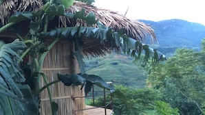 Uma construção com telhado de palha e plataforma de madeira, rodeada por vegetação exuberante e colinas ao fundo.