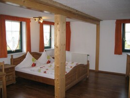 Uma cama com estrutura de madeira e colcha florida, duas janelas com cortinas laranja, uma mesinha de cabeceira de madeira e um radiador.