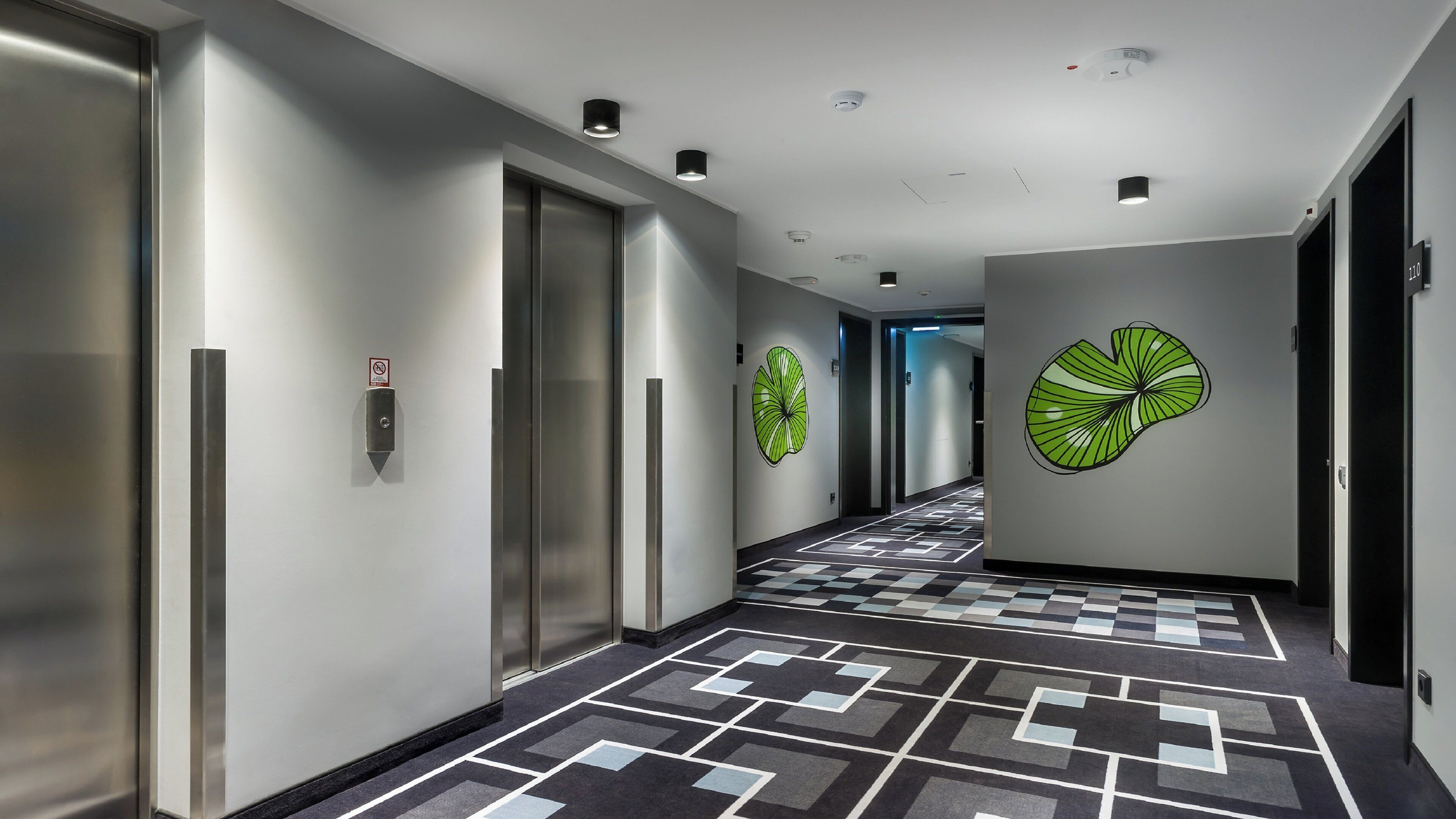 Corredor moderno de hotel com elevadores, carpete estampado e obras de arte nas paredes.