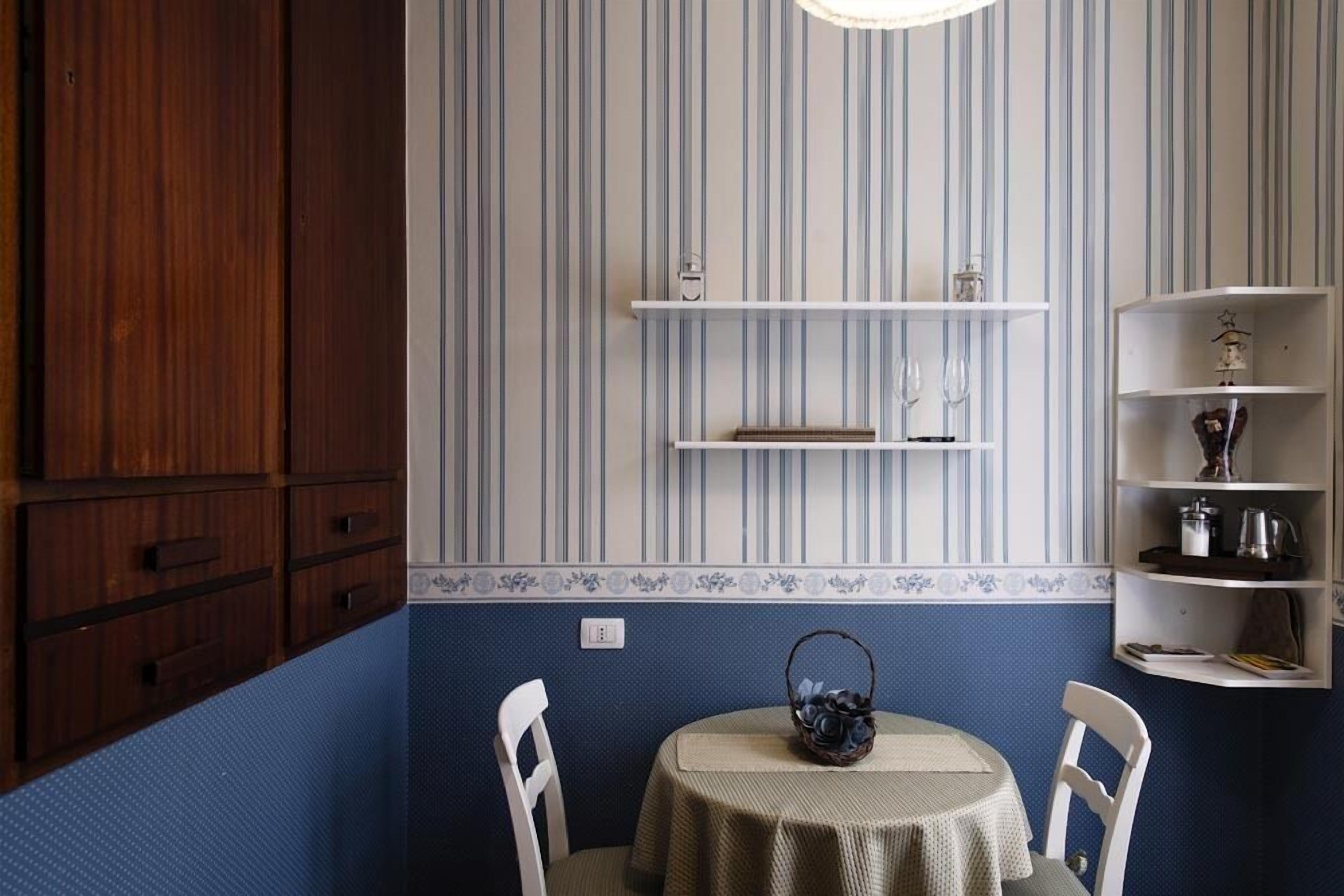 Uma área de refeições com uma mesa redonda, duas cadeiras, parede listrada e prateleiras com utensílios de cozinha.