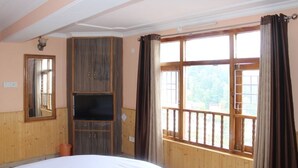 Quarto com cama, televisão, painéis de madeira, janela com cortinas e vista para o verde lá fora.