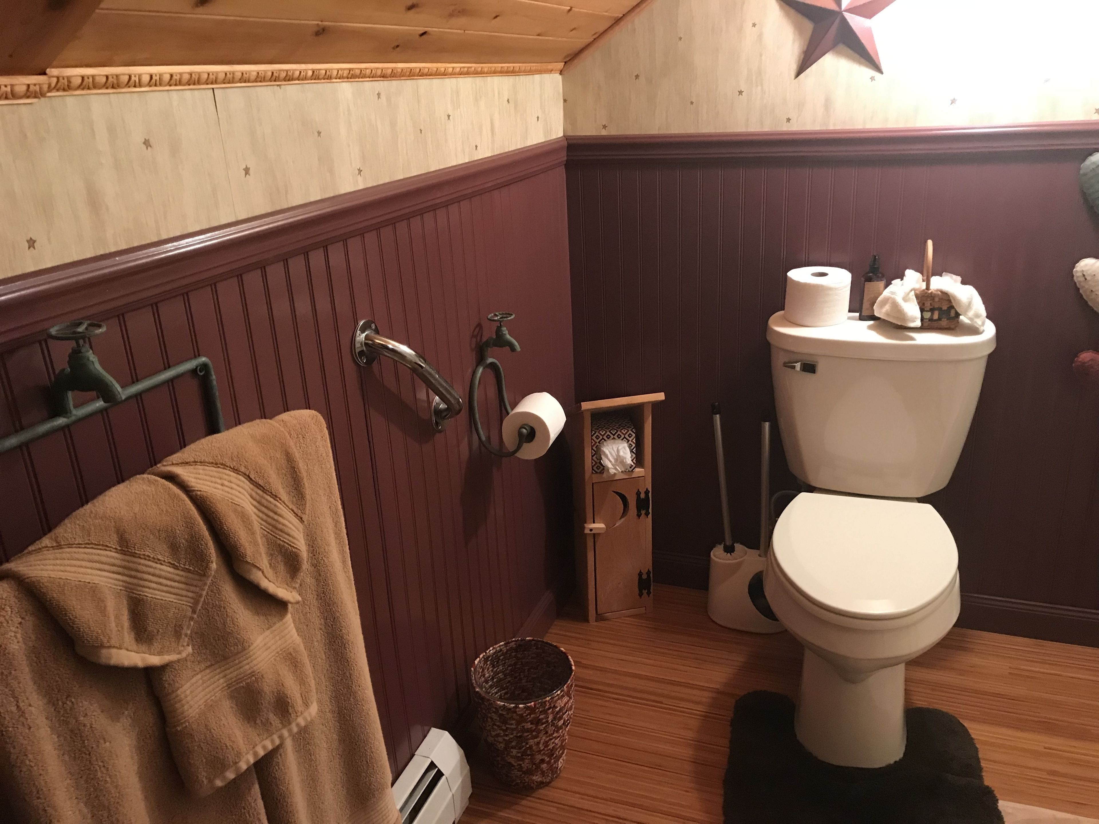 Uma casa de banho com soalho em madeira, sanita, porta-toalhas, dispensador de papel e vassoura.