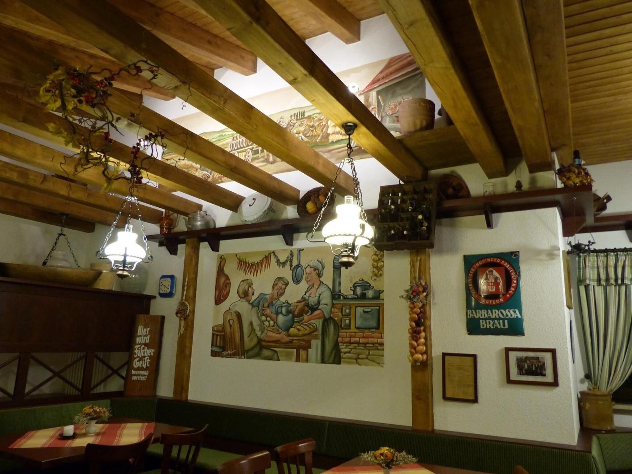Interior de restaurante com vigas de madeira, luminárias pendentes, um mural e uma placa com 'Barbarossa Brau'.