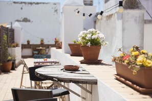 Terraço ao ar livre com mesa, cadeiras, plantas em vasos e luzes de Natal.