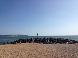 Uma praia de seixos com um molhe rochoso, céu azul límpido e mar calmo.