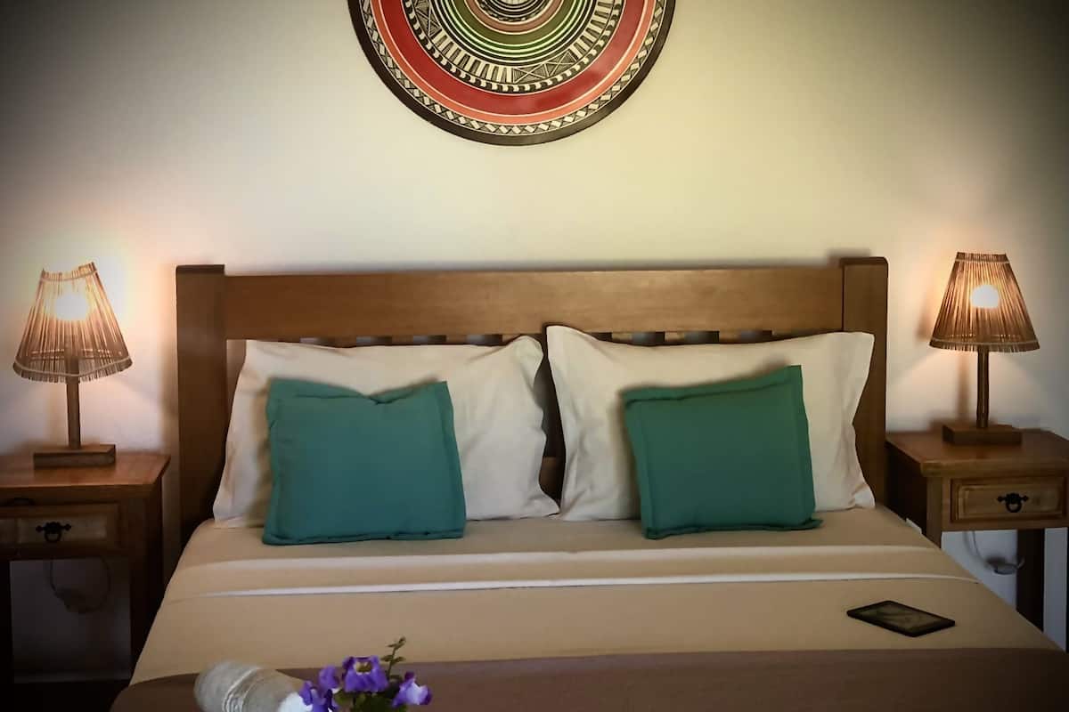 Cama bem arrumada com duas almofadas verdes, cabeceira de madeira e um enfeite decorativo na parede.