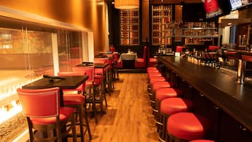 Área de bar com banquetas vermelhas, piso de madeira e uma parede de garrafas de vinho.