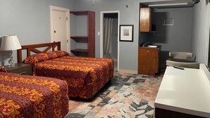 Quarto de hotel com duas camas, uma escrivaninha, uma cadeira, um ventilador de teto e um banheiro visível por uma porta aberta.