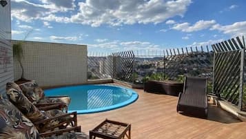Área de piscina no terraço com deck de madeira, espreguiçadeiras e vista para o horizonte da cidade.