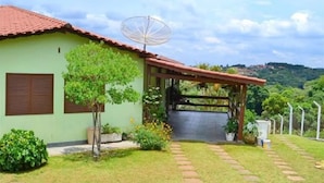 Uma casa com fachada verde, telhado telhado e uma pequena árvore, além de uma antena parabólica.