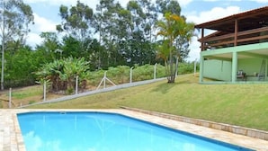 Uma piscina retangular com borda em mosaico, rodeada por um gramado e árvores.
