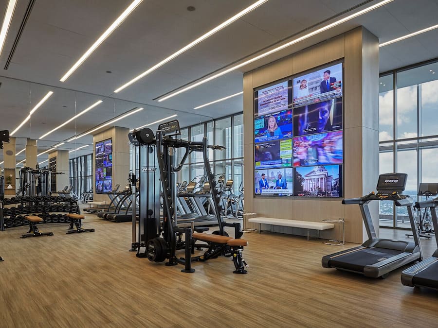 Uma academia moderna com equipamentos de cardio, máquinas de peso e uma parede de TV exibindo notícias.