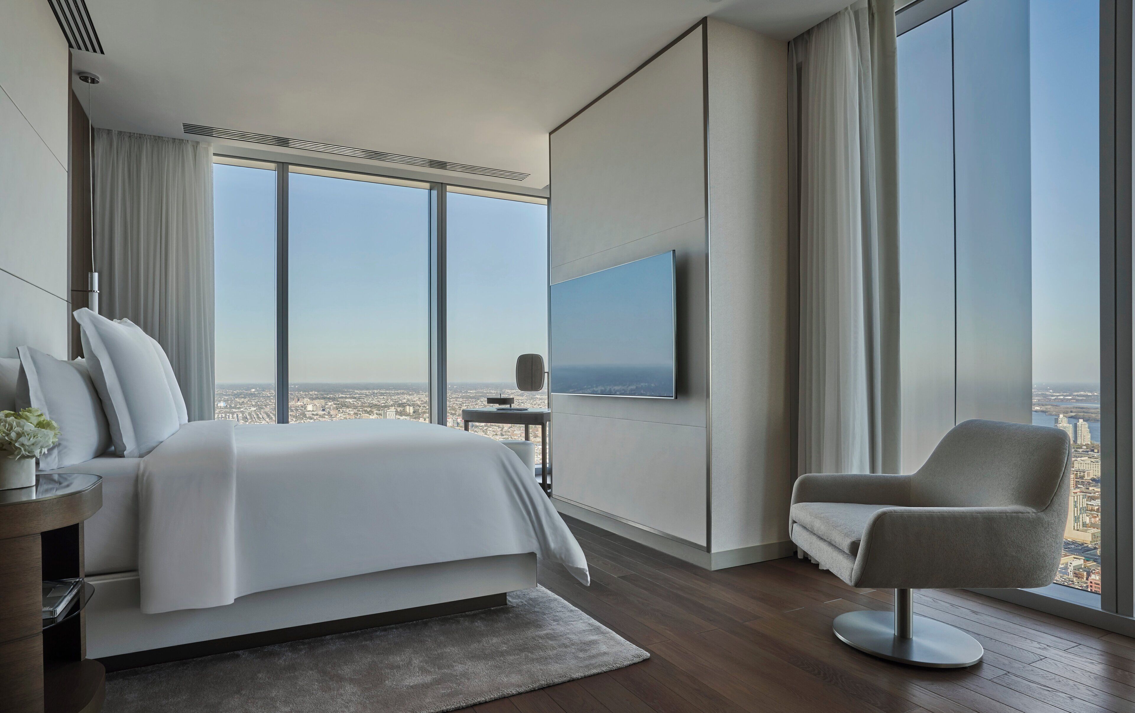 Quarto de hotel moderno com uma cama grande, televisão, cadeira e vista da cidade.