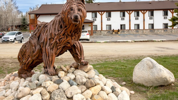 Uma escultura de urso de madeira sobre uma formação rochosa, com um edifício ao fundo.