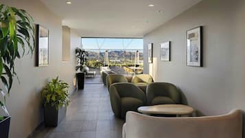 Um saguão de hotel moderno com vista para a cidade e montanhas. Há áreas de estar confortáveis e plantas em vasos.