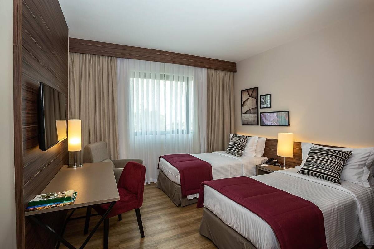 Quarto de hotel com duas camas, uma escrivaninha, uma cadeira, uma televisão e uma janela com cortinas.