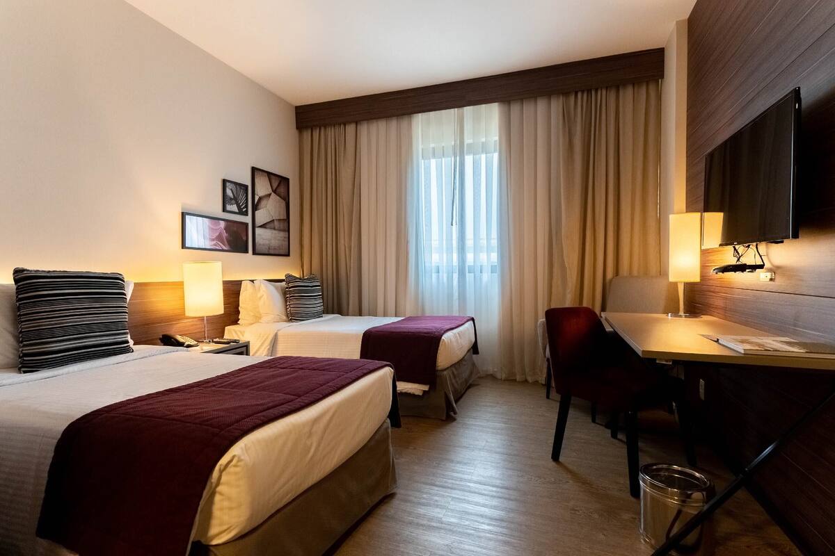 Quarto de hotel com duas camas, uma escrivaninha, uma cadeira, uma televisão e uma janela com cortinas.