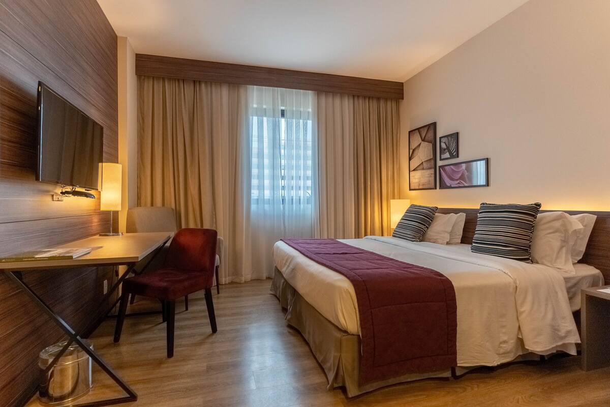 Quarto de hotel com cama, escrivaninha, cadeira, televisão na parede e janela com cortinas.