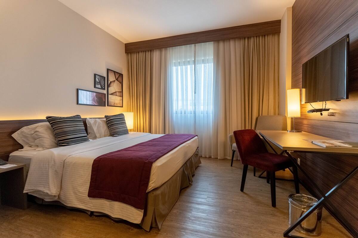 Quarto de hotel com cama, escrivaninha, cadeira e televisão na parede.