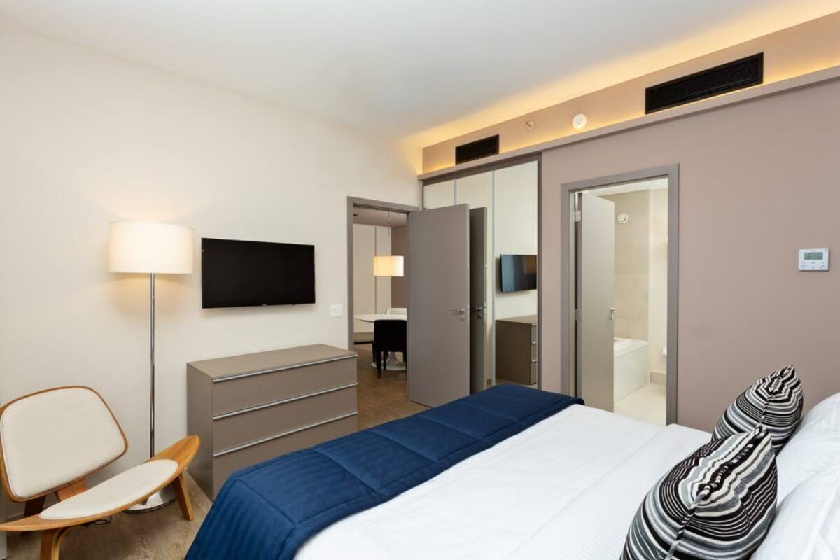 Quarto de hotel com cama, escrivaninha, cadeira, televisão e guarda-roupa.