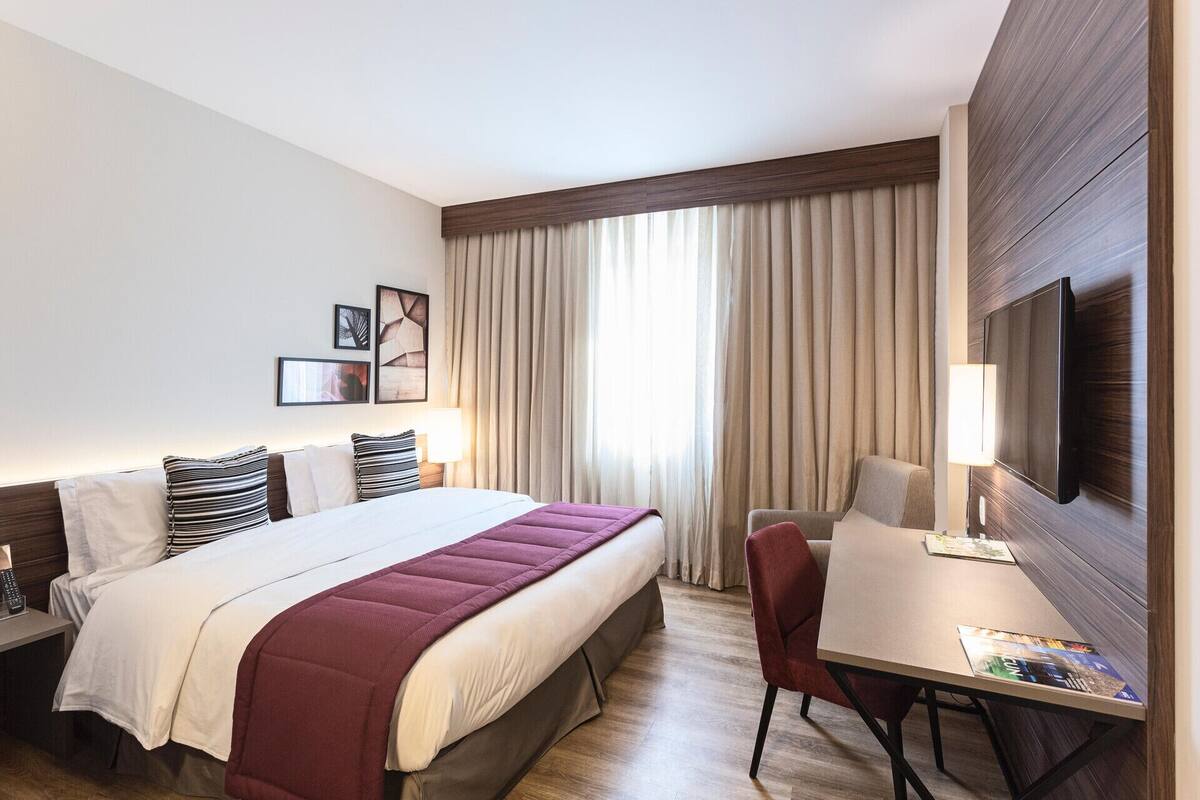Quarto de hotel com cama, mesa de cabeceira, cadeira, escrivaninha e televisão.