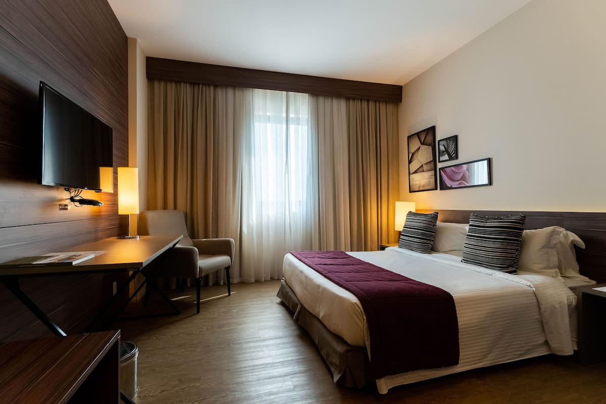 Quarto de hotel com uma cama, escrivaninha, cadeira, televisão e janela com cortinas.