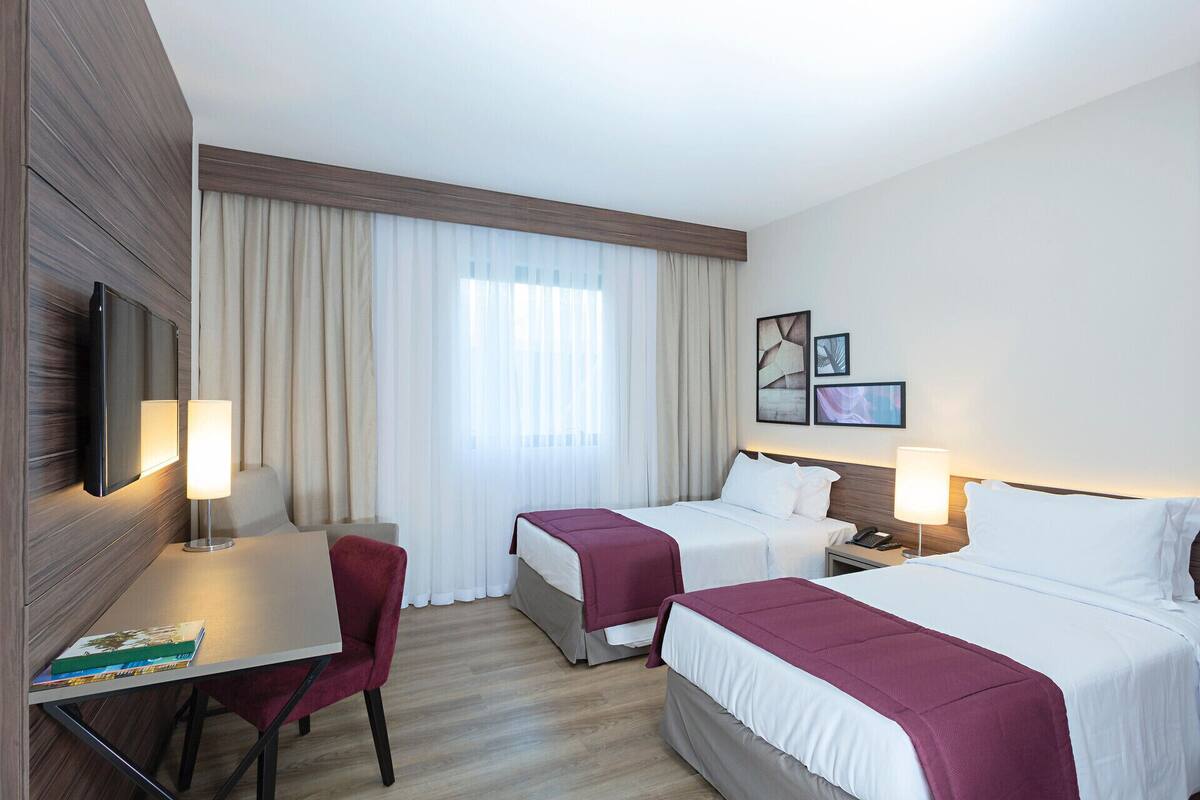 Quarto de hotel com duas camas, uma escrivaninha, uma cadeira, uma televisão na parede e uma janela com cortinas.
