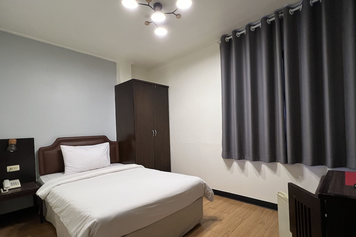 Quarto de hotel com cama, guarda-roupa, mesa de cabeceira, escrivaninha e janela com cortinas.