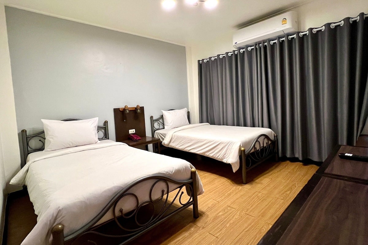 Quarto de hotel com duas camas, piso de madeira, mesa de cabeceira, luminária e janela com cortinas.