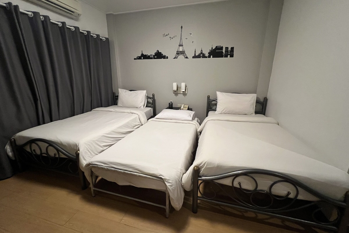 Quarto de hotel com duas camas de solteiro, ar condicionado fixado na parede e uma pintura urbana sobre as camas.
