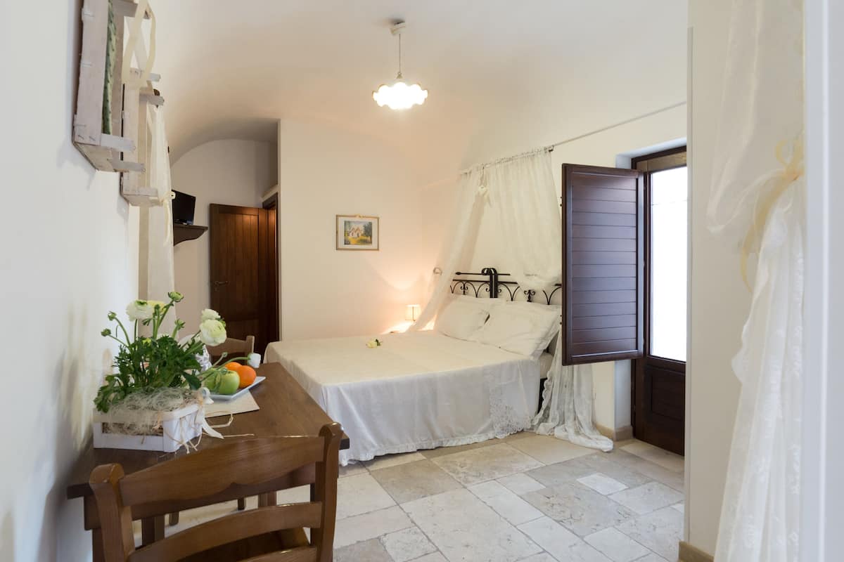 Quarto com cama, mesa de cabeceira com um vaso de flores, janela com cortinas e porta.