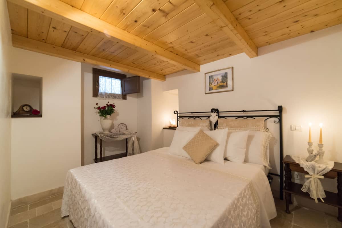 Um quarto com teto de madeira, cama com roupa de cama branca, uma mesinha com um vaso de flores e um relógio fixado na parede.