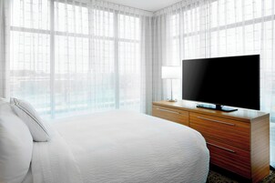 Quarto de hotel com uma cama grande, penteadeira de madeira, TV de tela plana e cortinas translúcidas.