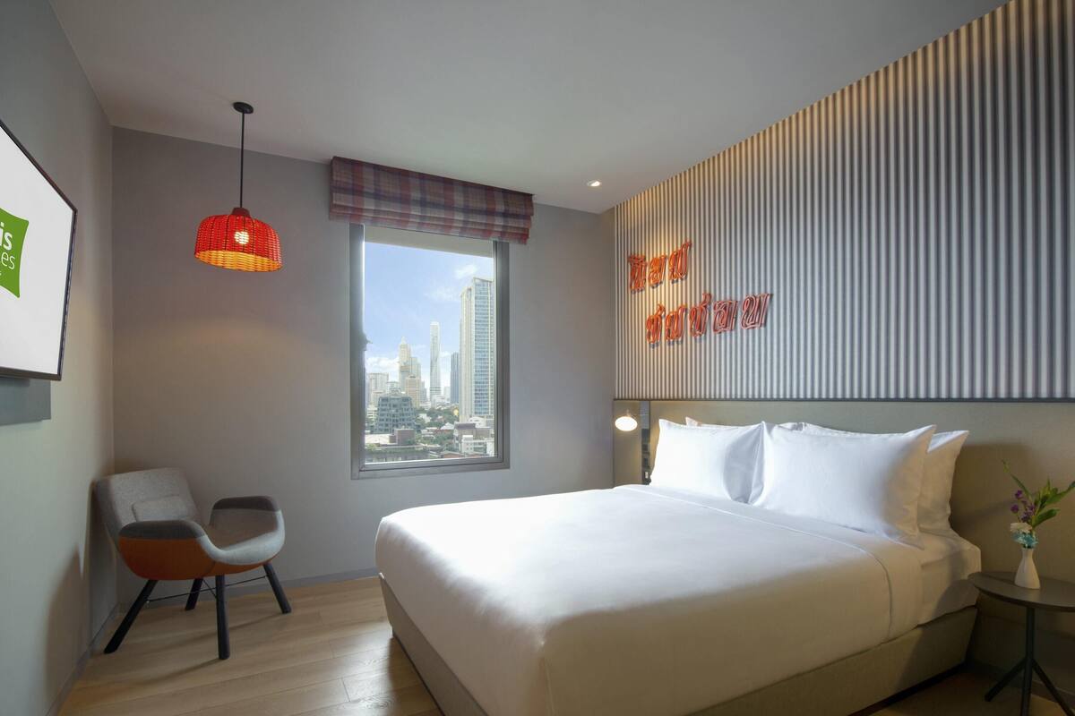 Quarto de hotel moderno com uma cama grande, vista para a cidade, luminária pendente vermelha e uma pequena mesa com um vaso.
