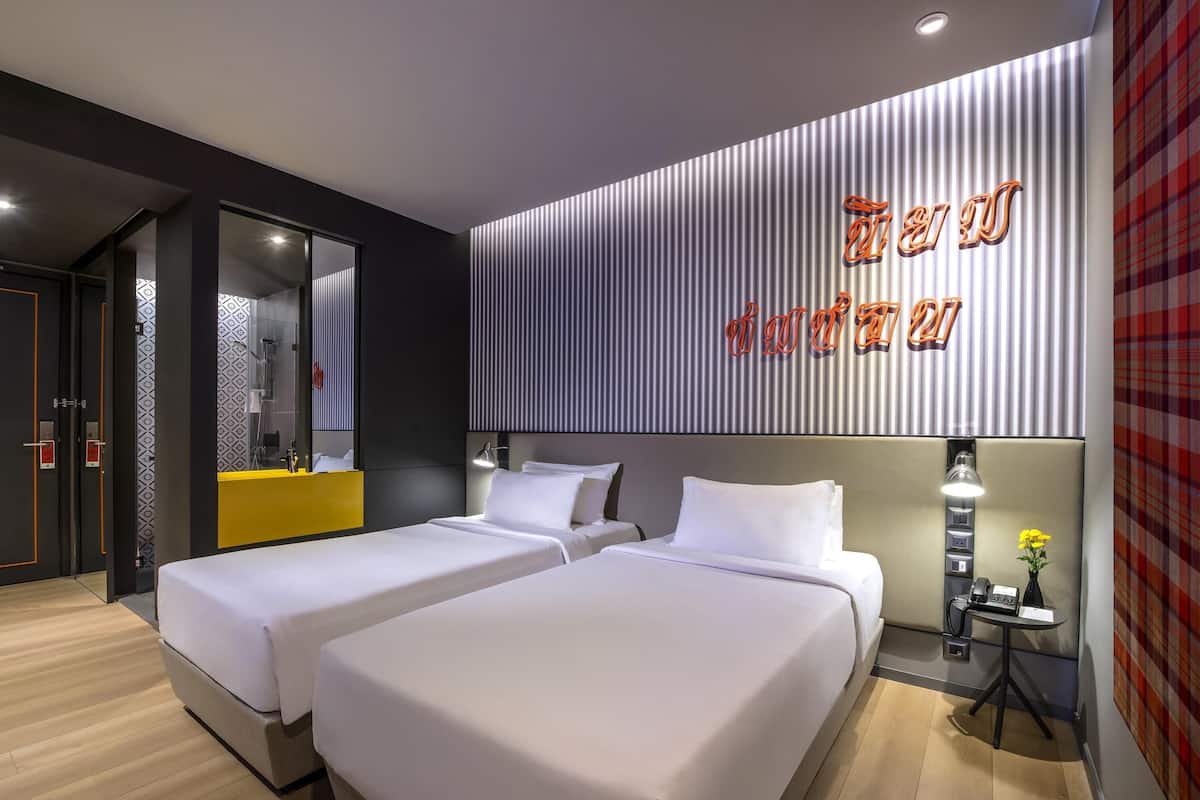 Quarto de hotel com duas camas, um espelho, mesa de cabeceira com abajur e iluminação na parede.