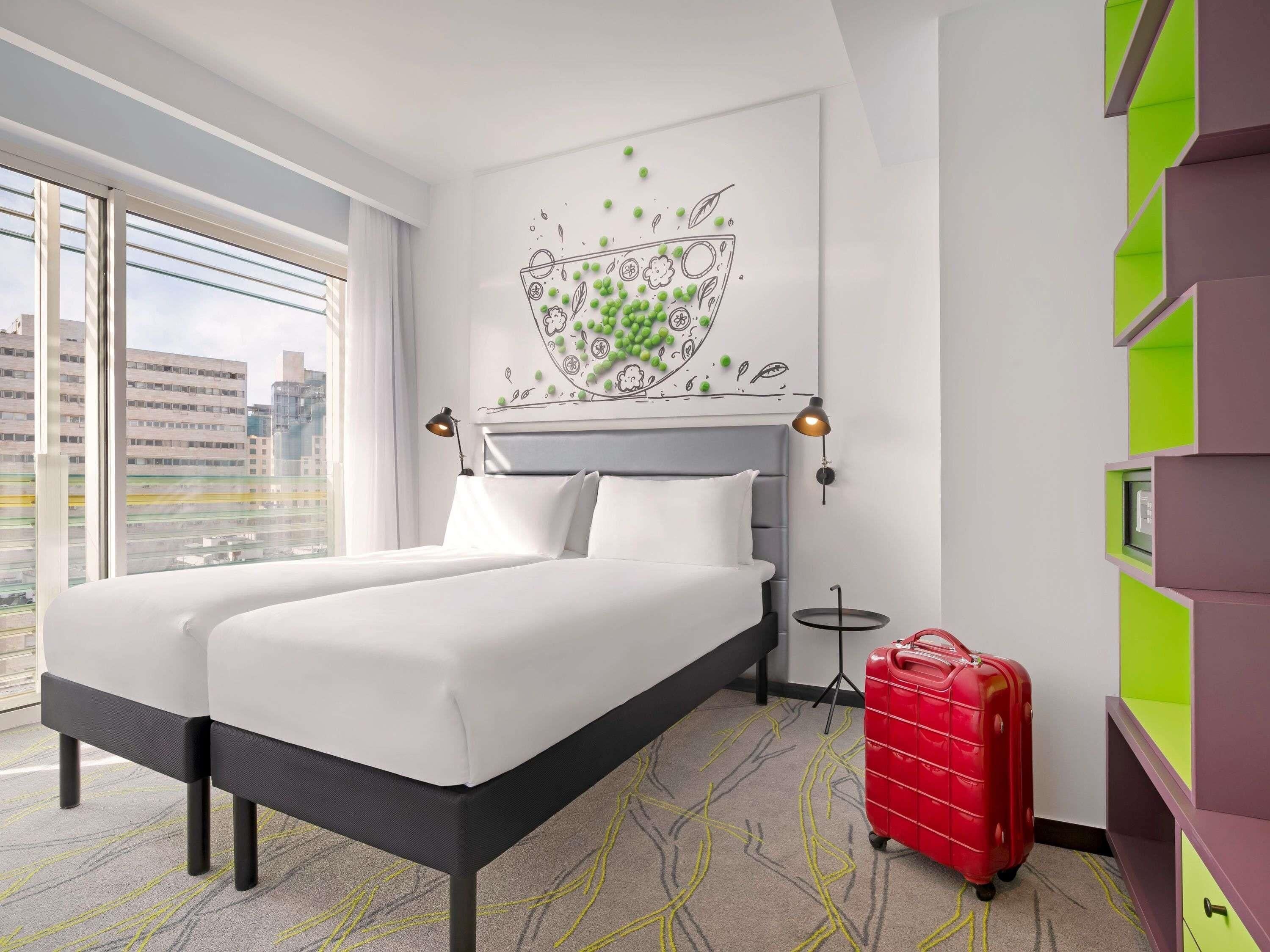 Quarto de hotel moderno com uma cama grande, vista para a cidade, arte única na parede e uma mala vermelha.