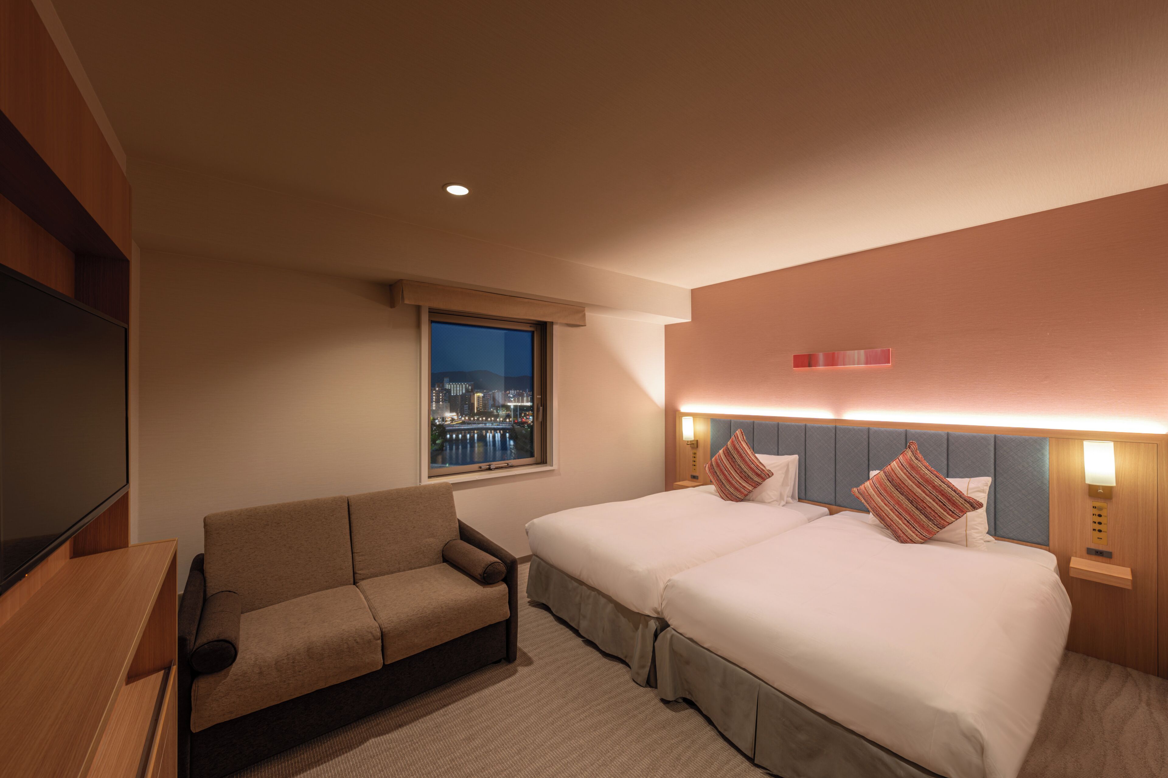 Quarto de hotel com televisão, sofá, duas camas, janela com vista para a cidade e mesa de cabeceira com abajur.