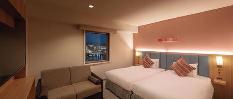 Quarto de hotel com televisão, sofá, duas camas, janela com vista para a cidade e mesa de cabeceira com abajur.