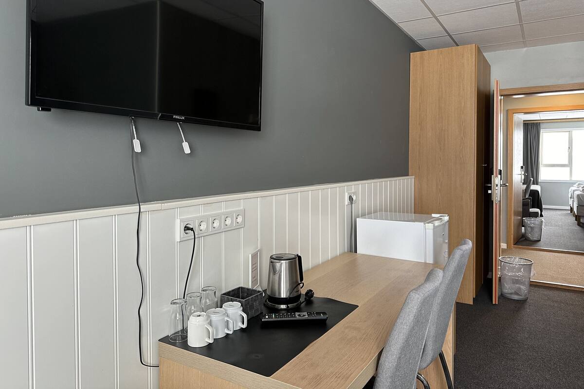 Quarto de hotel com televisão de tela plana presa à parede, mesa de madeira com cafeteira, canecas e controle remoto, e vista do quarto adjacente por uma porta aberta.