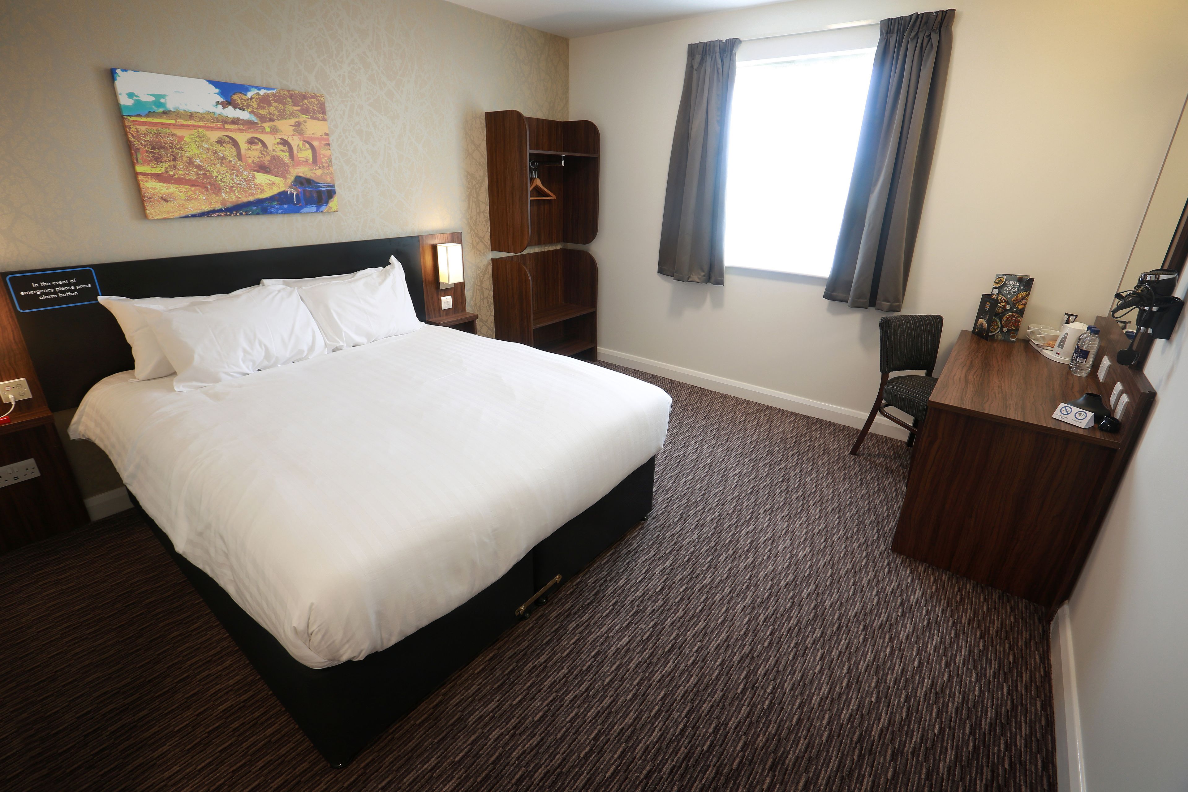 Quarto de hotel com uma cama grande, um guarda-roupa de madeira, uma escrivaninha com cadeira e uma janela com cortinas.