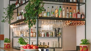Uma área de bar com balcão de mosaico verde, dois banquetas pretas, prateleiras com garrafas e copos, e plantas em vasos.