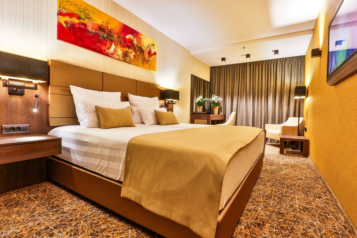 Quarto de hotel com uma cama grande, uma mesa de cabeceira, uma escrivaninha, uma cadeira, uma TV e um quadro na parede.