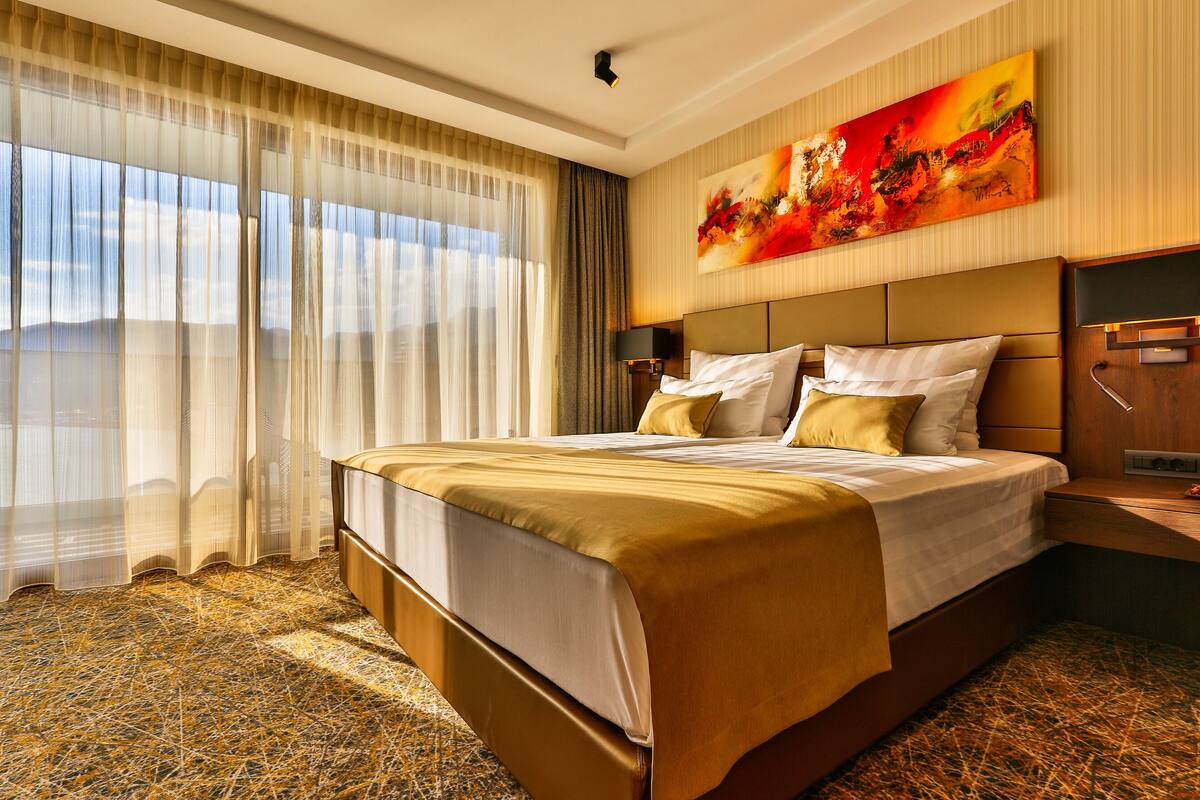 Quarto de hotel com uma cama grande, cabeceira, mesa de cabeceira com uma luminária e um quadro na parede.