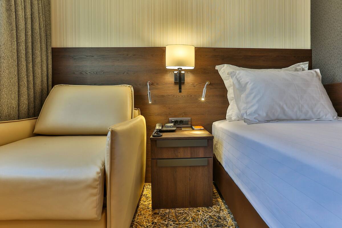Quarto de hotel com uma cama, uma mesa de cabeceira com abajur, uma cadeira e uma pequena escrivaninha com telefone e um livro.