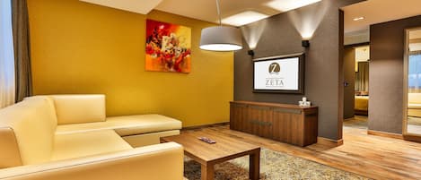 Quarto de hotel moderno com sofá, mesa de centro de madeira, televisão e um quadro na parede.